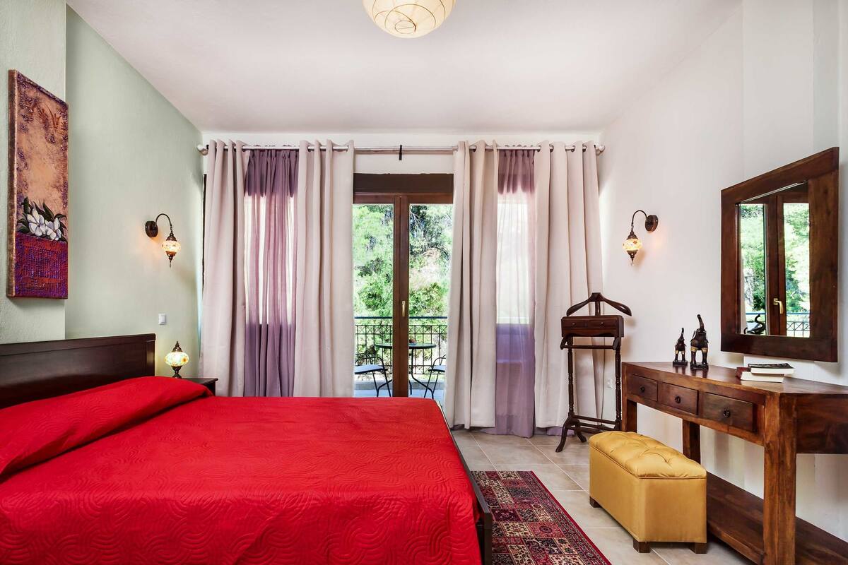 Quarto com colcha vermelha, mesa de cabeceira de madeira, penteadeira e um banco. Há duas arandelas, um quadro e vista para o exterior pela janela.