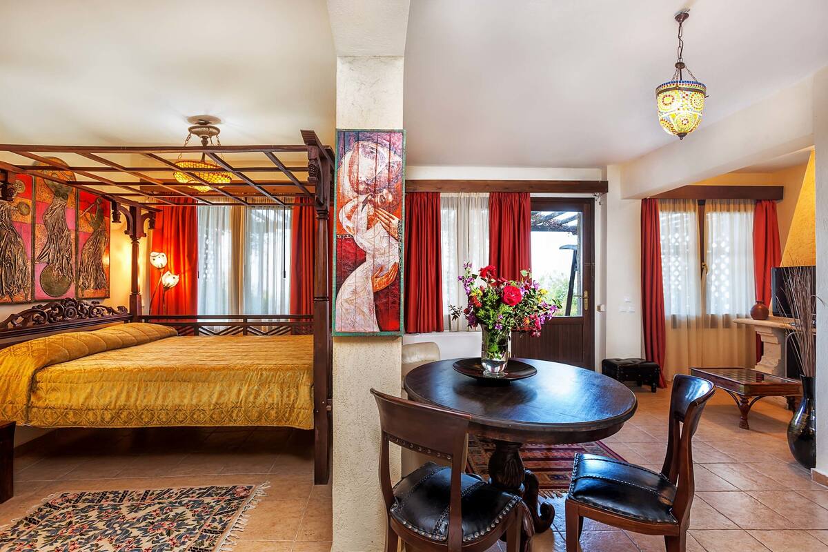 Um quarto com uma cama, uma mesa de jantar com cadeiras, um candelabro e uma pintura grande na parede.