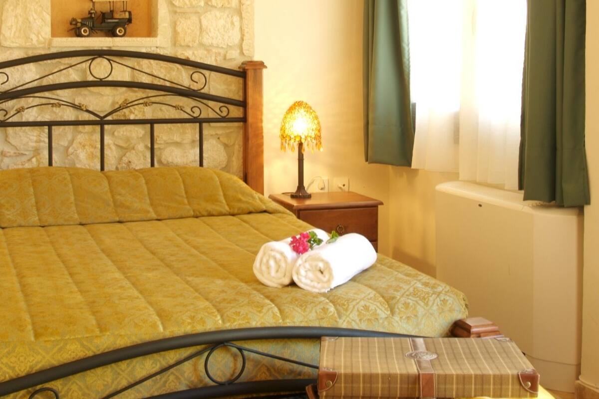 Uma cama com um edredom, uma parede de pedra, uma janela com cortinas e um abajur.