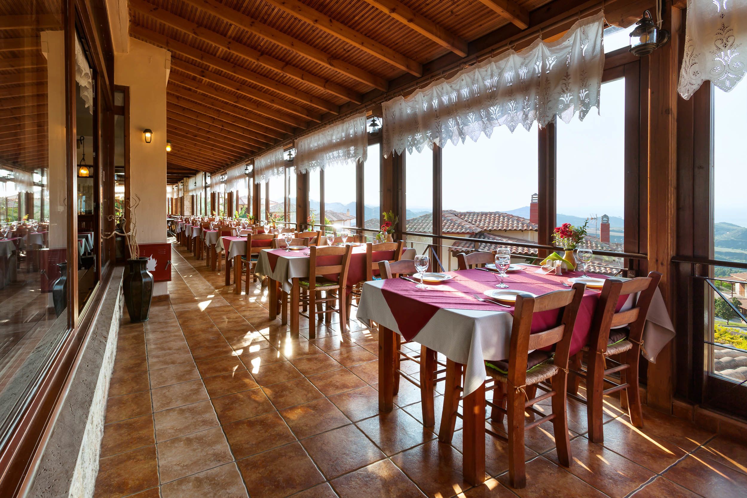 Restaurante com vigas de madeira e janelas amplas, oferecendo vista para as montanhas. Mesas dispostas com toalhas brancas e guardanapos vermelhos.