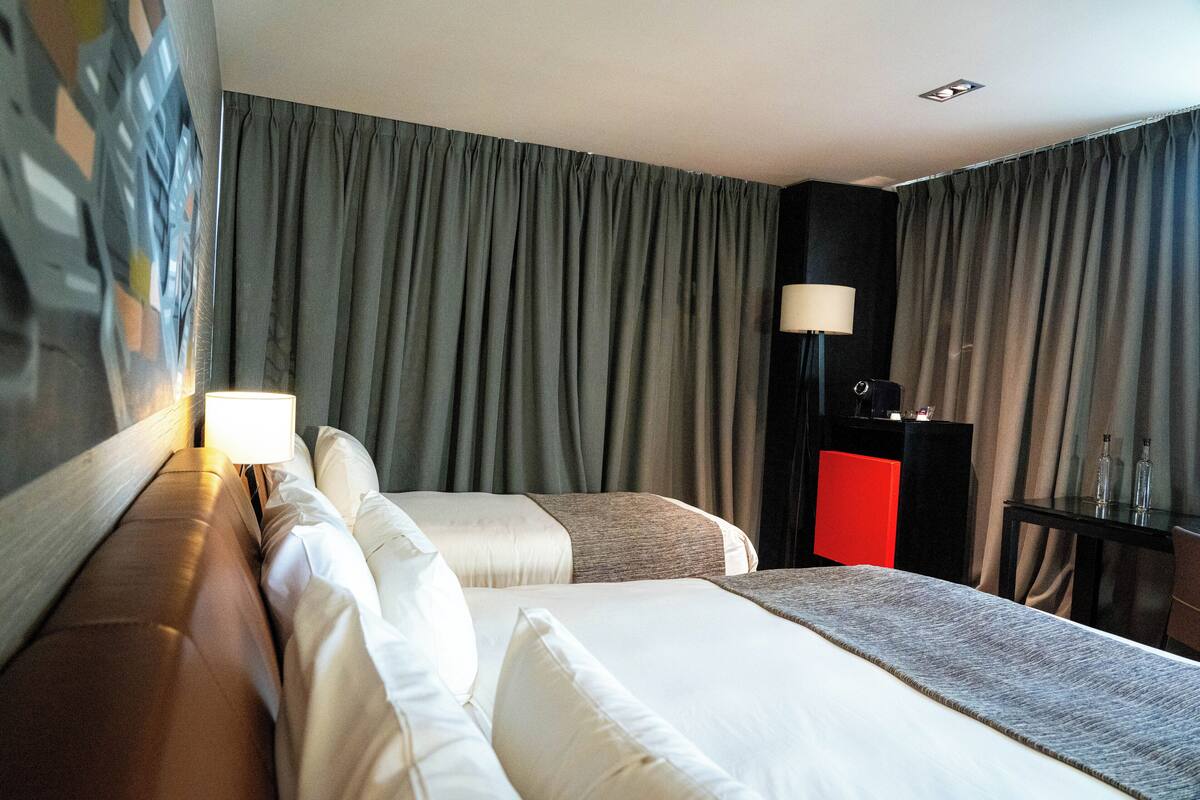 Quarto de hotel com duas camas, uma escrivaninha, uma lâmpada e uma cadeira vermelha.