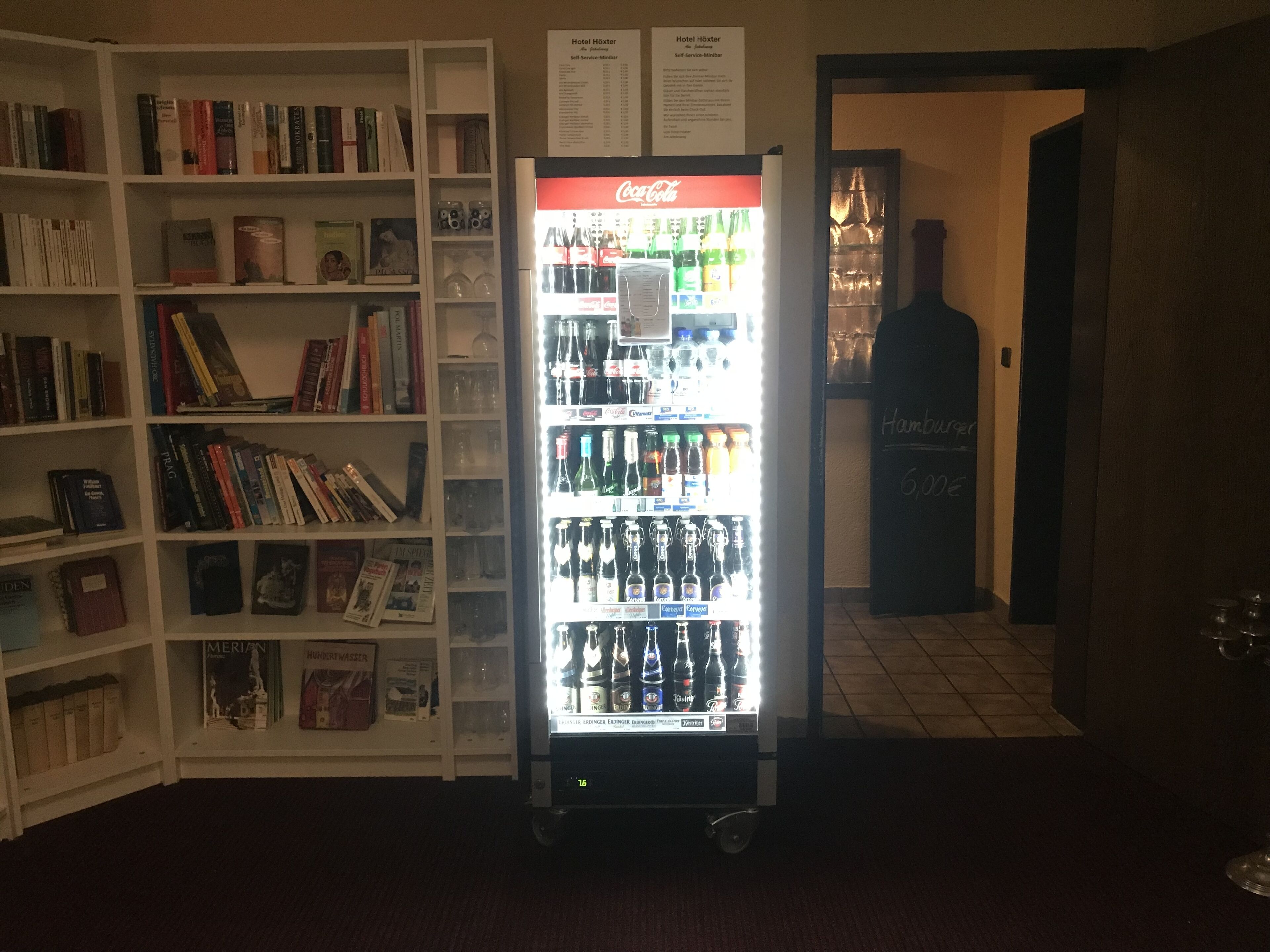 Uma geladeira Coca-Cola repleta de diversas bebidas, cercada por estantes cheias de livros e revistas.