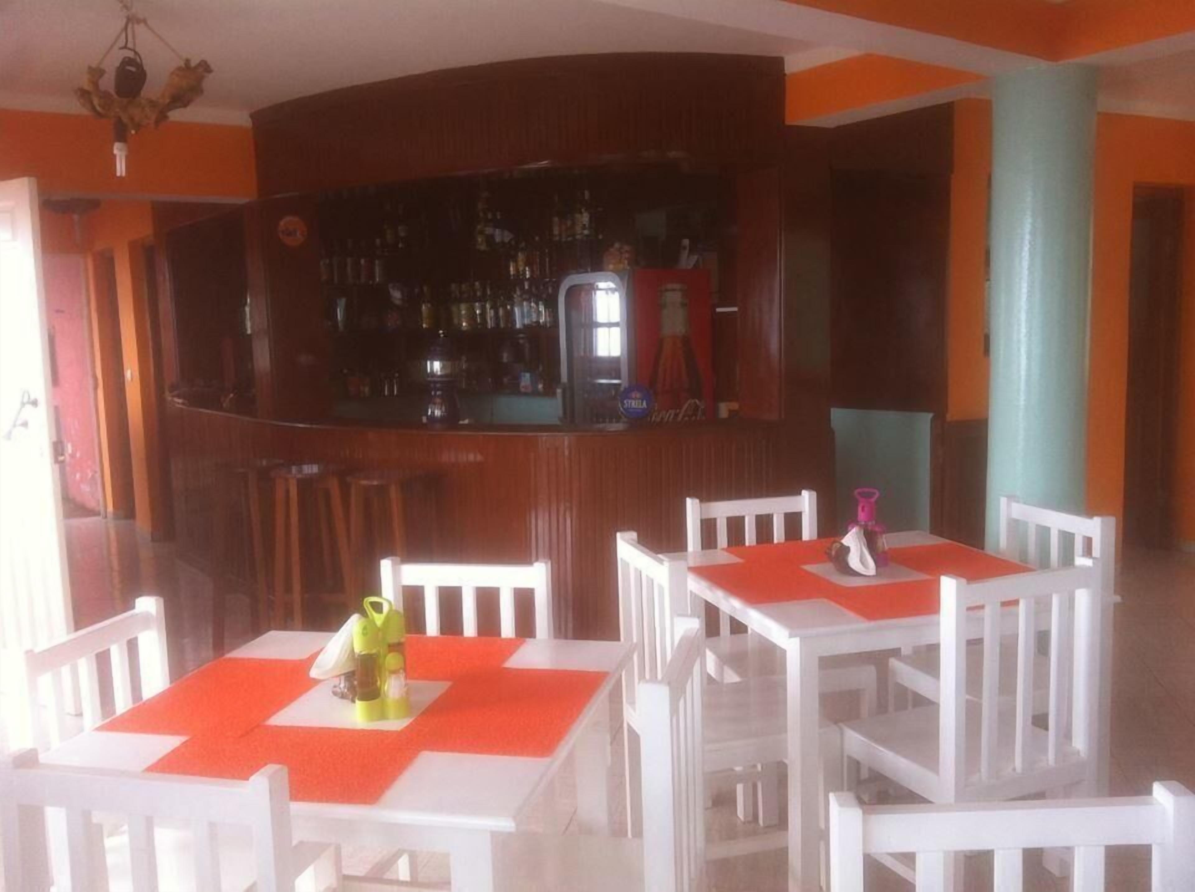 Restaurante com cadeiras e mesas brancas, área de bar com prateleiras de garrafas e um balcão de madeira curvado.