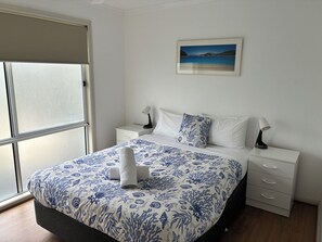 Quarto com cama, mesa de cabeceira, gaveta, janela e quadro na parede.