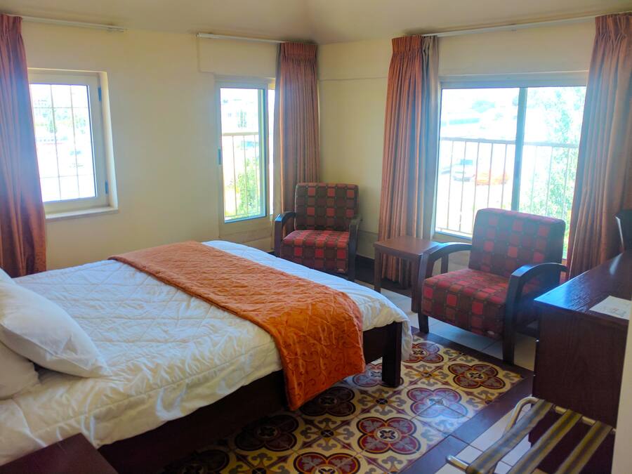 Quarto de hotel com cama, duas cadeiras, uma mesinha e uma janela com cortinas.