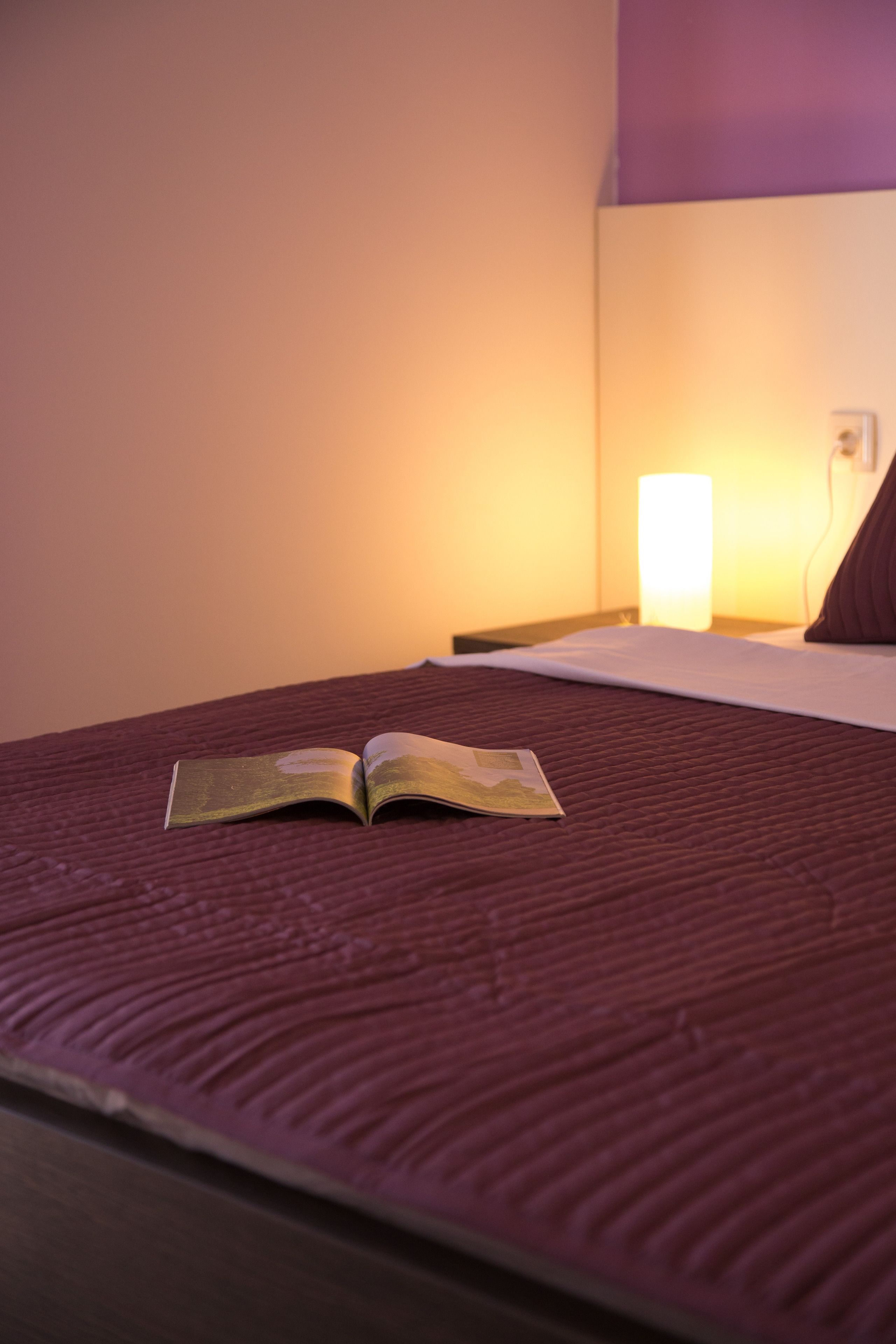 Uma cama com um livro, uma luminária de cabeceira e um travesseiro.