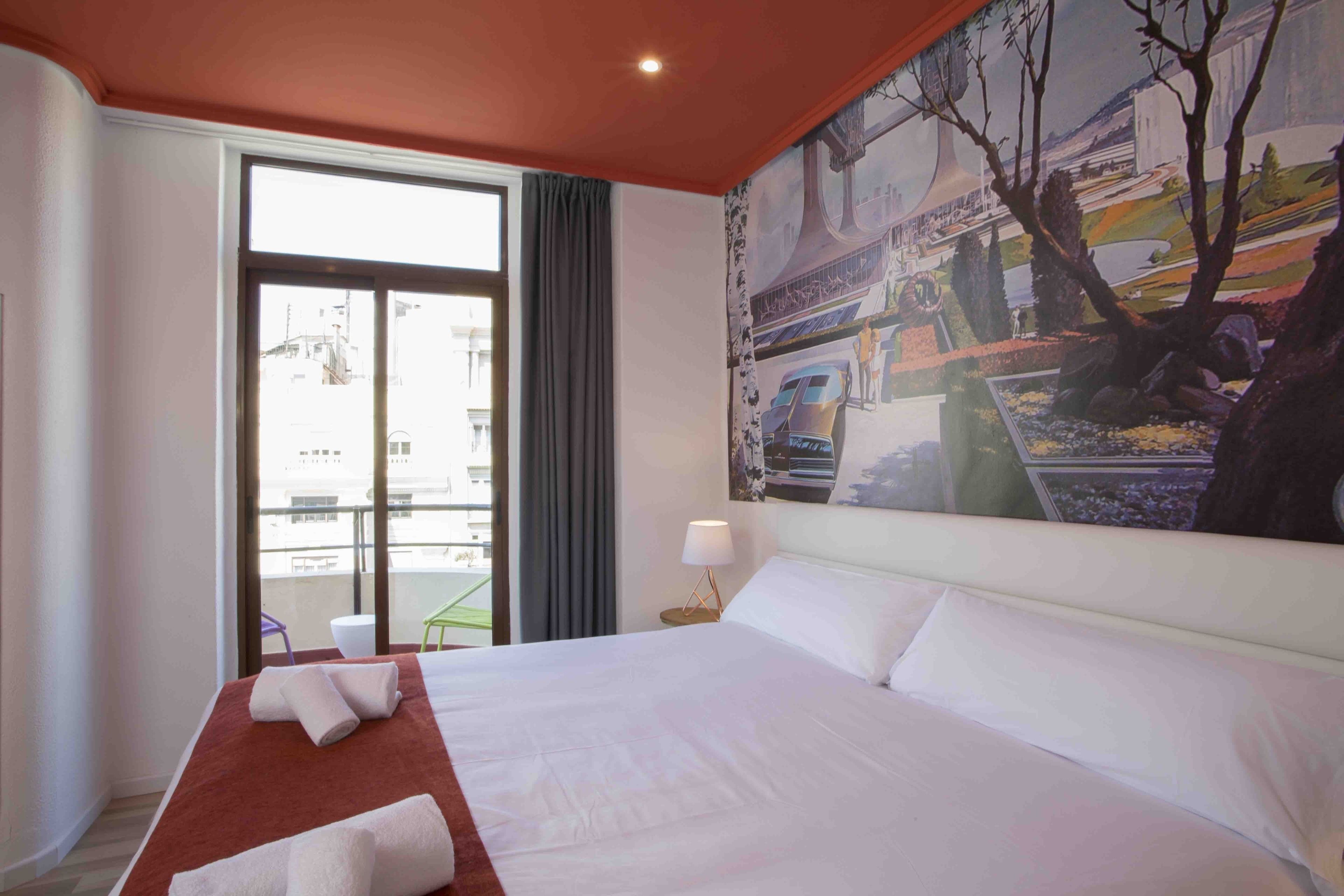 Quarto de hotel com uma cama grande, janela com vista para edifícios, um quadro na parede e uma mesinha com abajur.