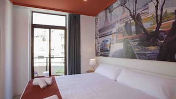 Quarto de hotel com uma cama grande, janela com vista para edifícios, um quadro na parede e uma mesinha com abajur.