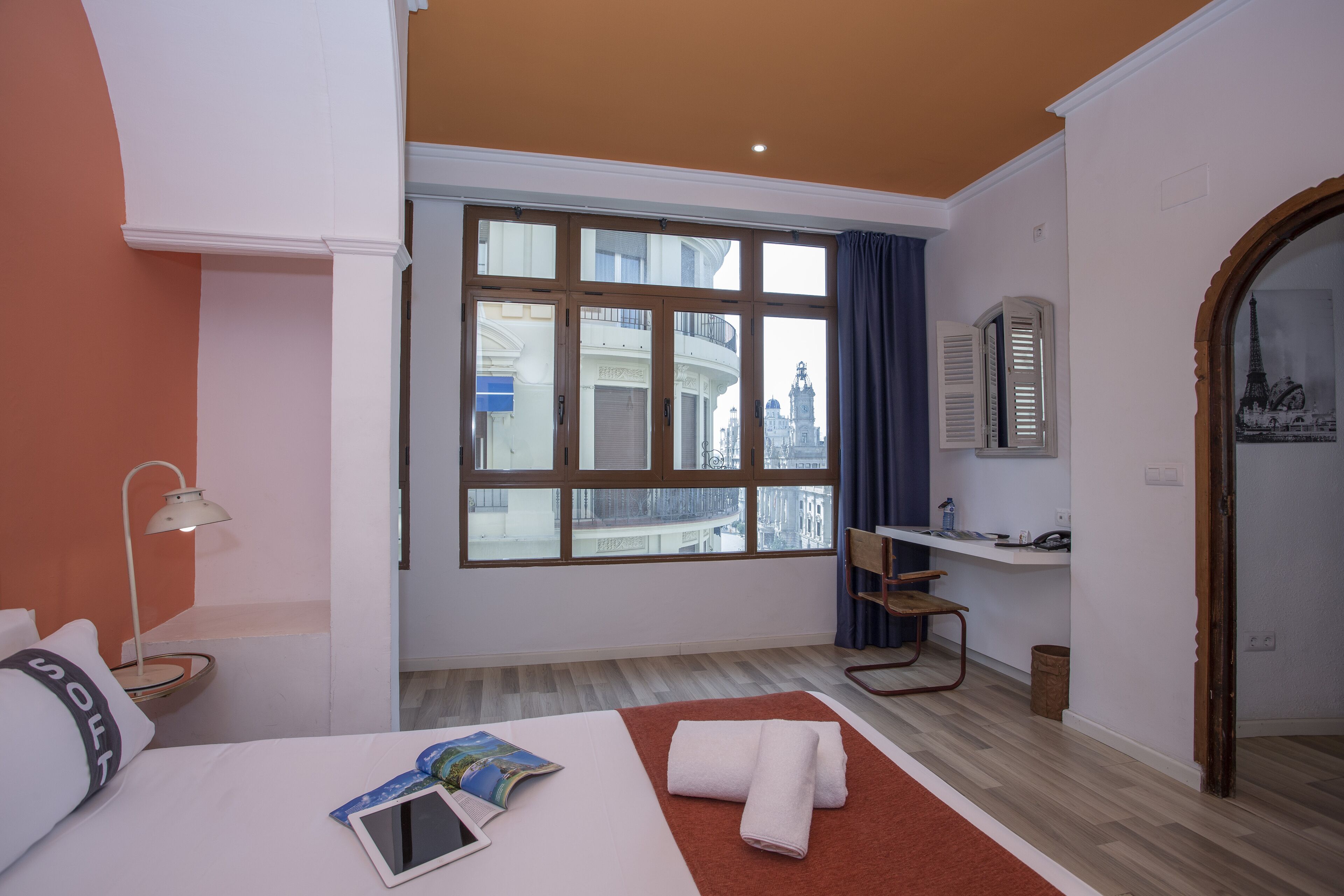 Quarto de hotel com cama, escrivaninha, cadeira, janela com vista para edifícios e espelho com imagem da Torre Eiffel.