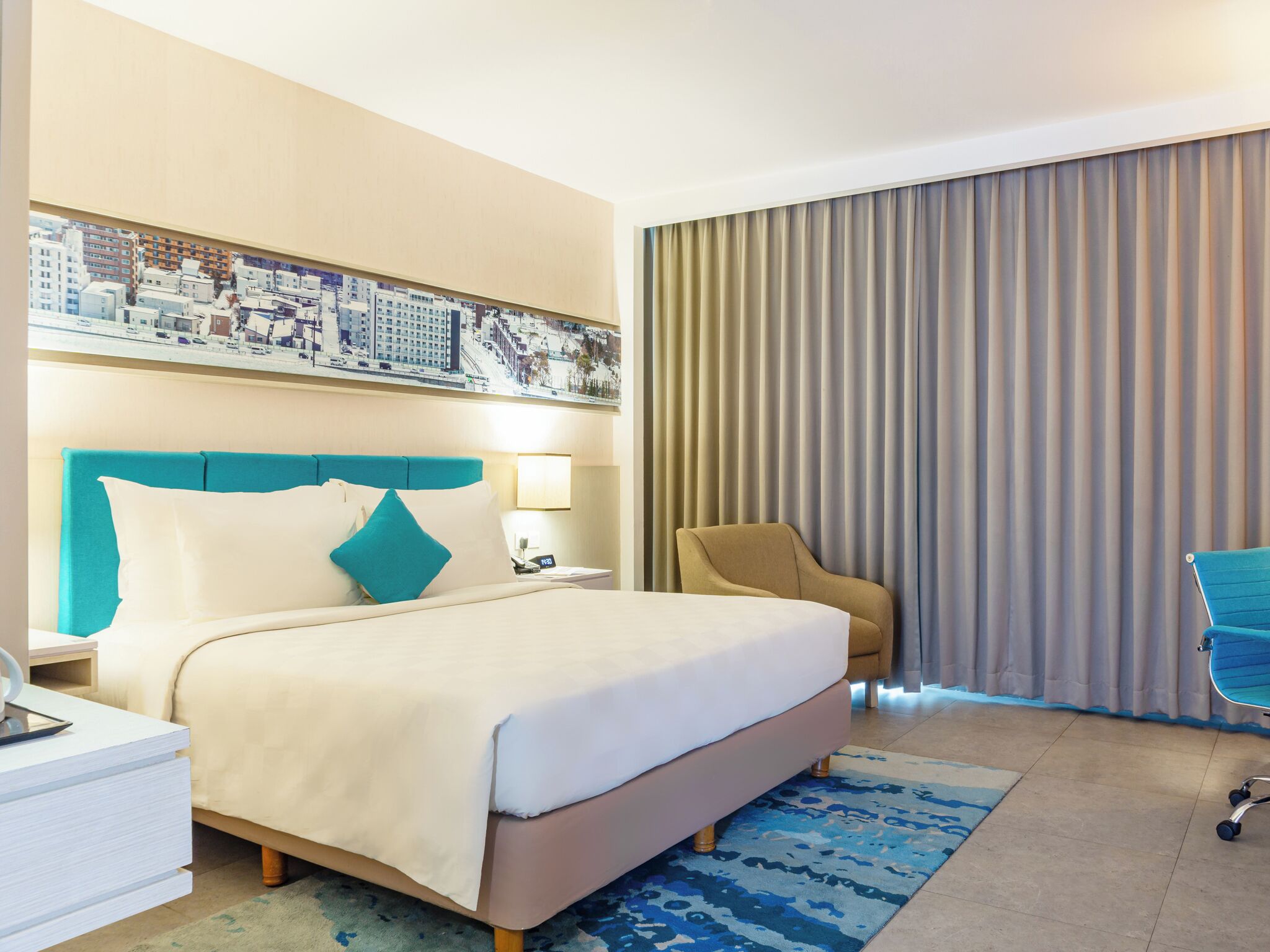Quarto de hotel com cama, uma cadeira, escrivaninha com abajur e vista para a cidade.