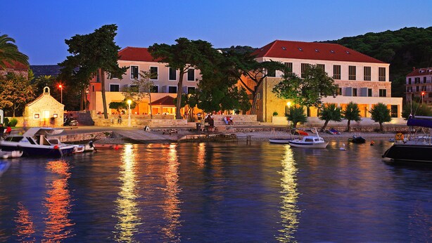 Hotel na orla com vários edifícios, um cais com barcos e um rio calmo refletindo luzes.
