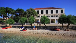 Um edifício com fachada de pedra e telhado de telhas vermelhas, cercado por árvores e uma área de praia com pedriscos e caiaques.