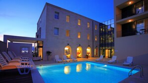 Uma piscina com espreguiçadeiras, um edifício com várias janelas e varandas, e um céu claro.