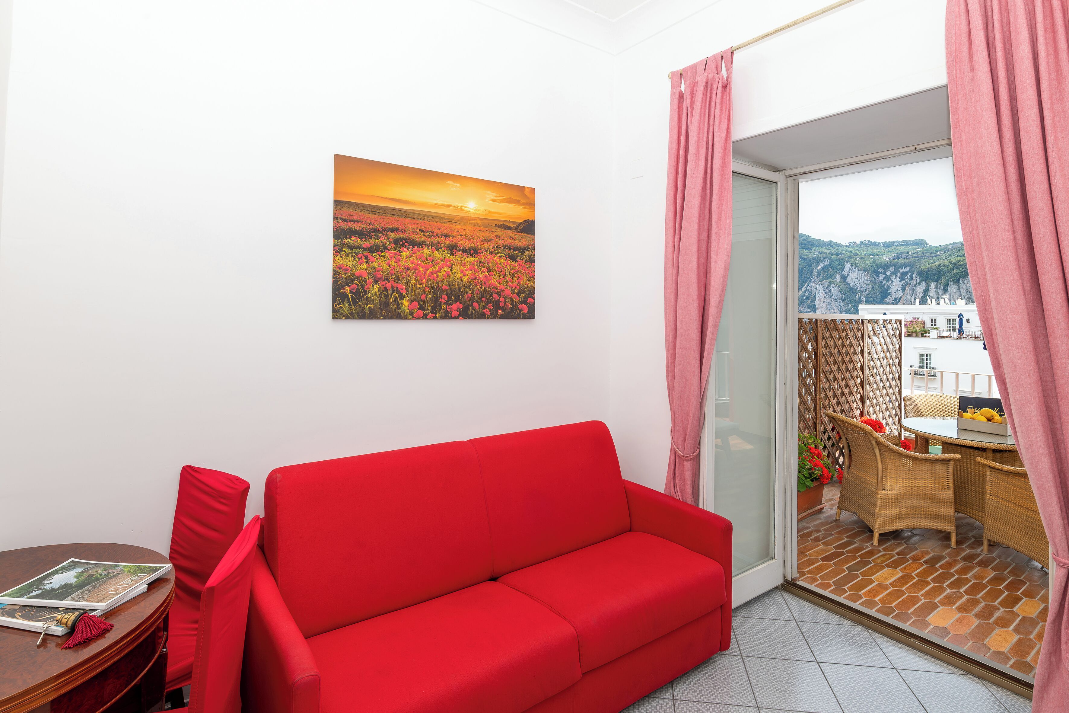 Um sofá vermelho, uma mesa com uma revista, e uma varanda com móveis de vime e vista para uma paisagem montanhosa.