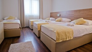 Quarto de hotel com três camas, cada uma com cabeceira bege, roupa de cama branca e almofadas amarelas. O chão é de madeira e há uma janela grande com cortinas.