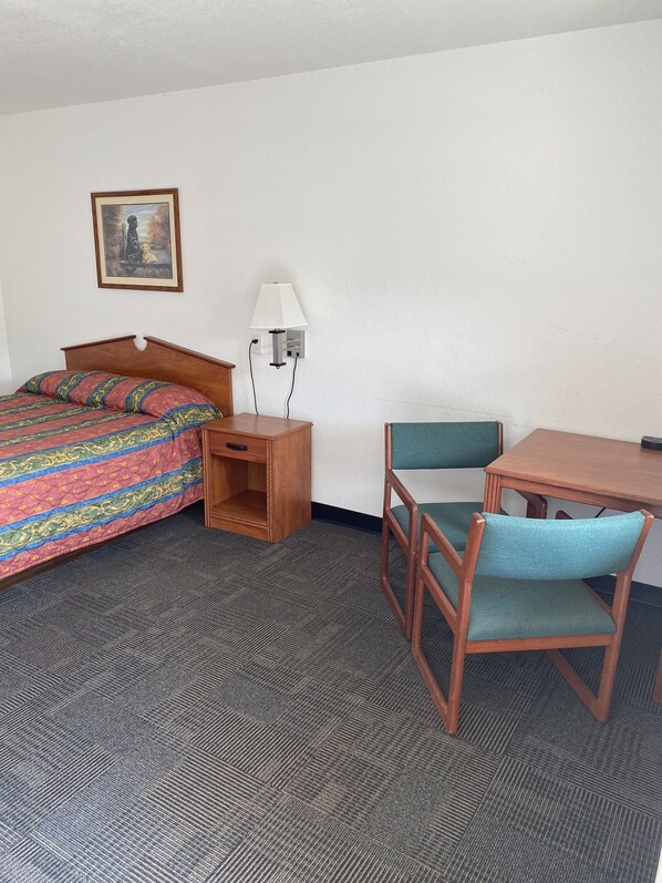 Quarto de hotel com cama, escrivaninha, cadeira, luminária e um quadro na parede.