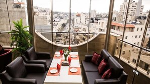 Uma sala de jantar com uma mesa posta para dois, dois sofás pretos e uma janela grande com vista para a cidade.