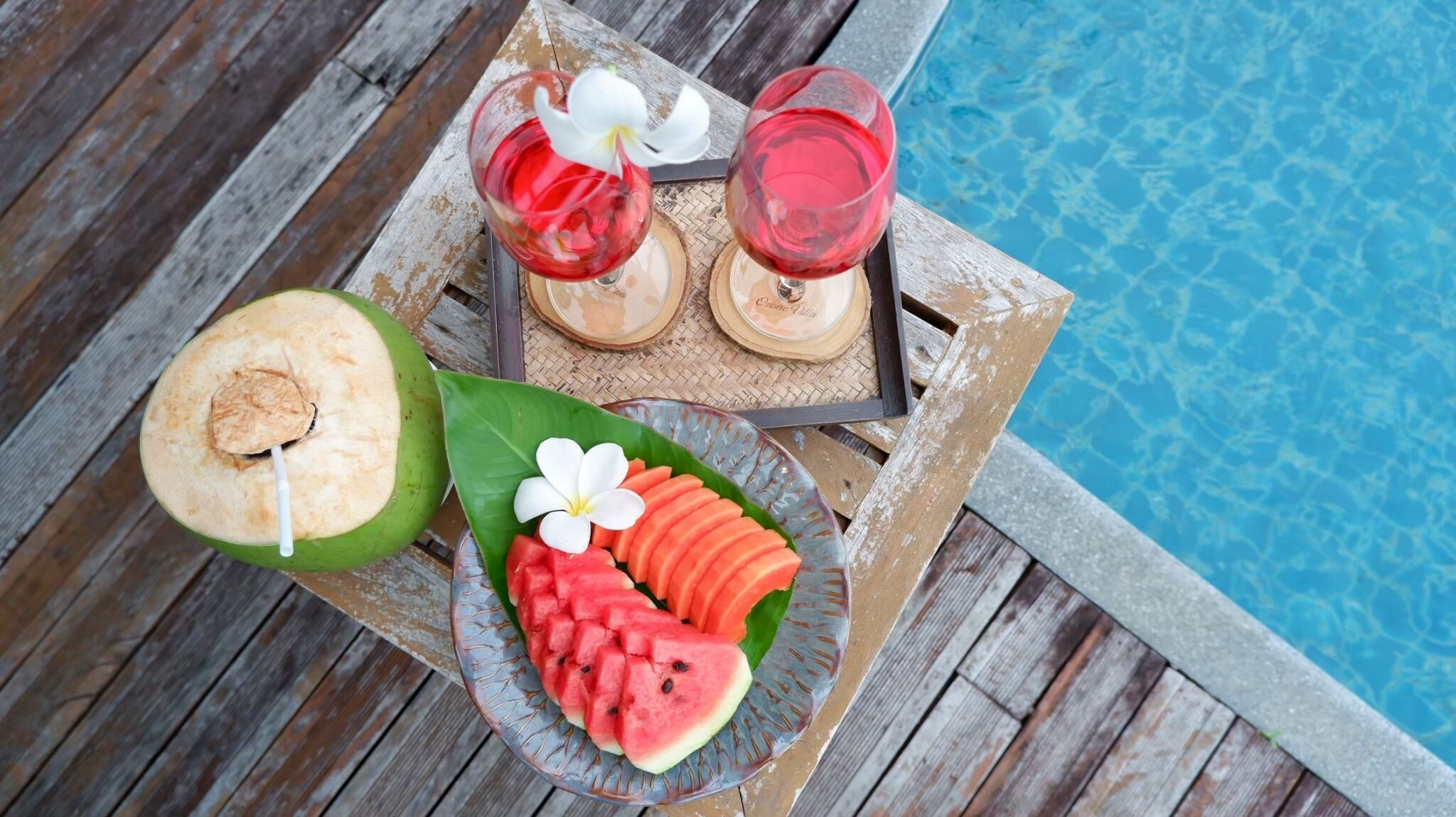 Um deck de madeira com piscina, uma travessa de melancia, um coco e duas bebidas vermelhas decoradas com flores.