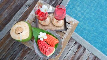 Um deck de madeira com piscina, uma travessa de melancia, um coco e duas bebidas vermelhas decoradas com flores.