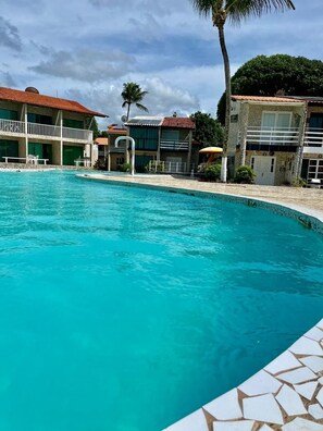 Uma piscina com água azul cristalina, cercada por edifícios com varandas e uma palmeira.