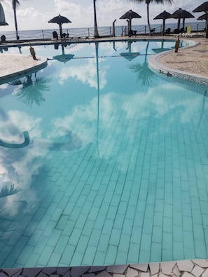 Piscina límpida com bordas azulejadas, rodeada por espreguiçadeiras e guarda-sóis, com vista para o oceano.