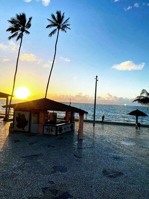 Um quiosque à beira-mar com piso de azulejos, palmeiras e vista para o mar ao pôr do sol.