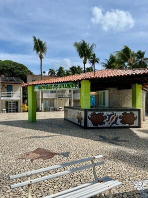 Quiosque chamado 'Kiosque do Village' com telhado de azulejos e um banco de mosaico na frente.