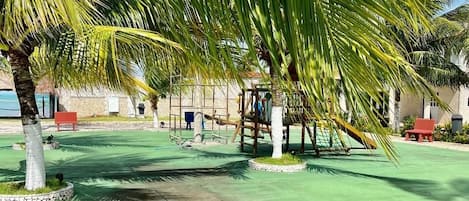 Um parquinho com escorrega e balanços, rodeado por palmeiras e um piso verde.