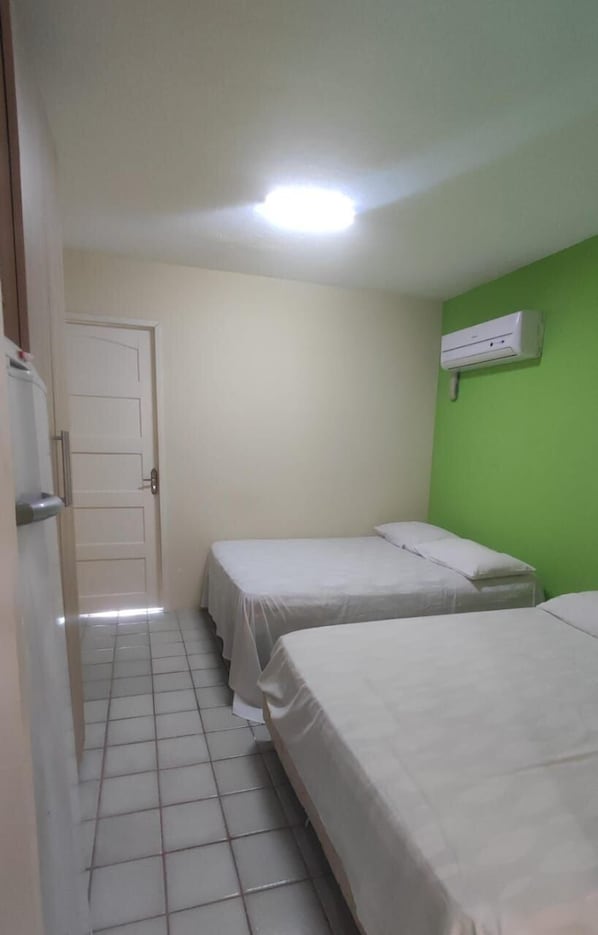 Quarto pequeno e limpo, com duas camas, uma porta, ar condicionado fixado na parede e piso em azulejos.