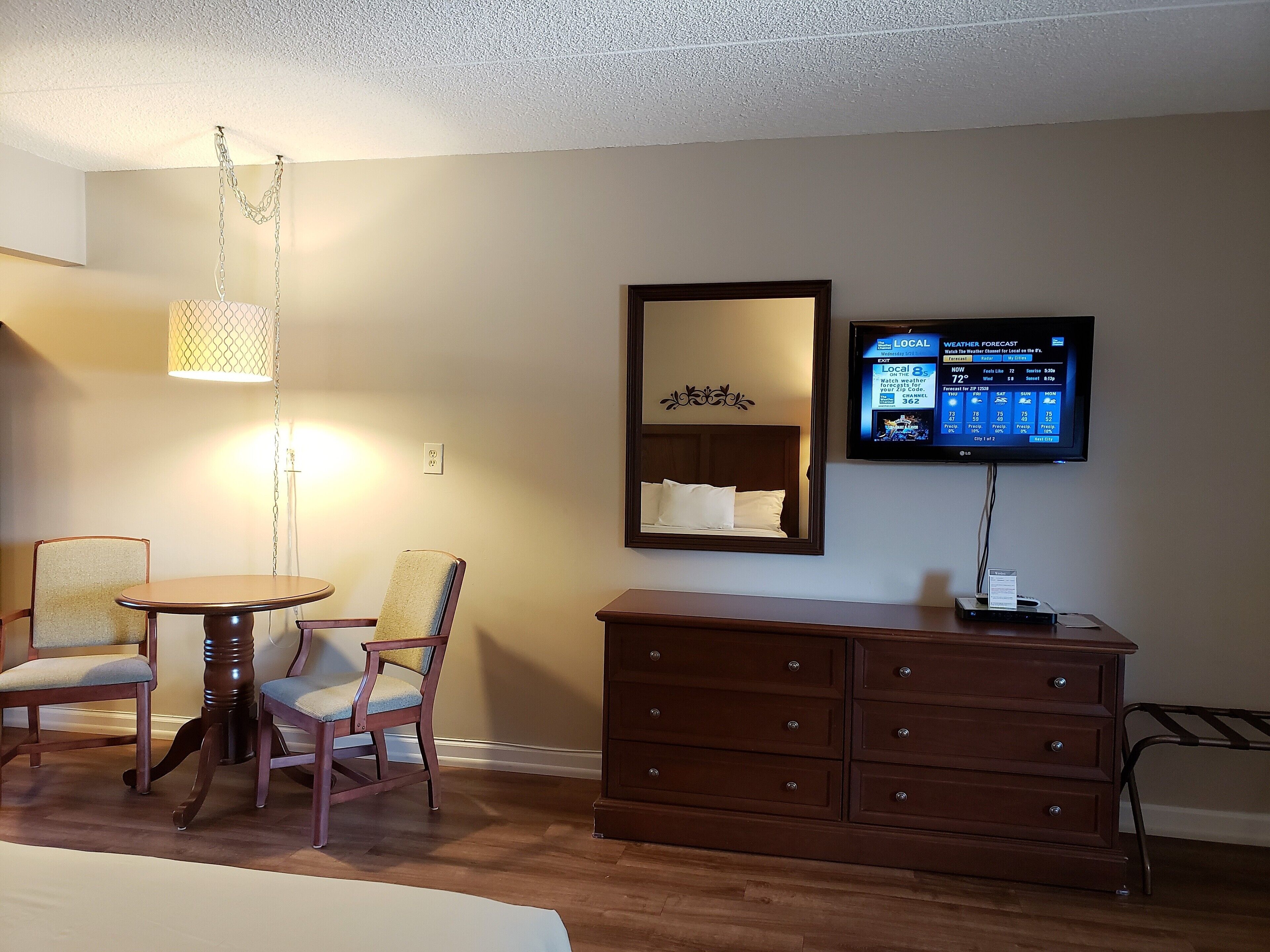 Quarto de hotel com cama, cómoda de madeira, televisão exibindo a previsão do tempo e área de jantar com cadeiras e mesa.