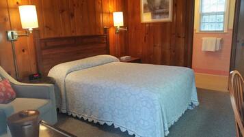 Um quarto com cabeceira de madeira, duas luminárias de apoio, uma cama com colcha floral, uma mesa de cabeceira com relógio e uma janela com vista para árvores.