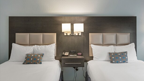 Quarto de hotel com duas camas, cada uma com travesseiro, uma mesa de cabeceira com luminária, e um criado-mudo com telefone e relógio.