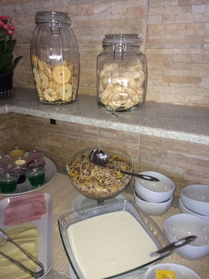 Disposição de buffet com diversos alimentos, incluindo uma tigela de cereal, um prato com bananas fatiadas e potes com biscoitos e outros lanches.