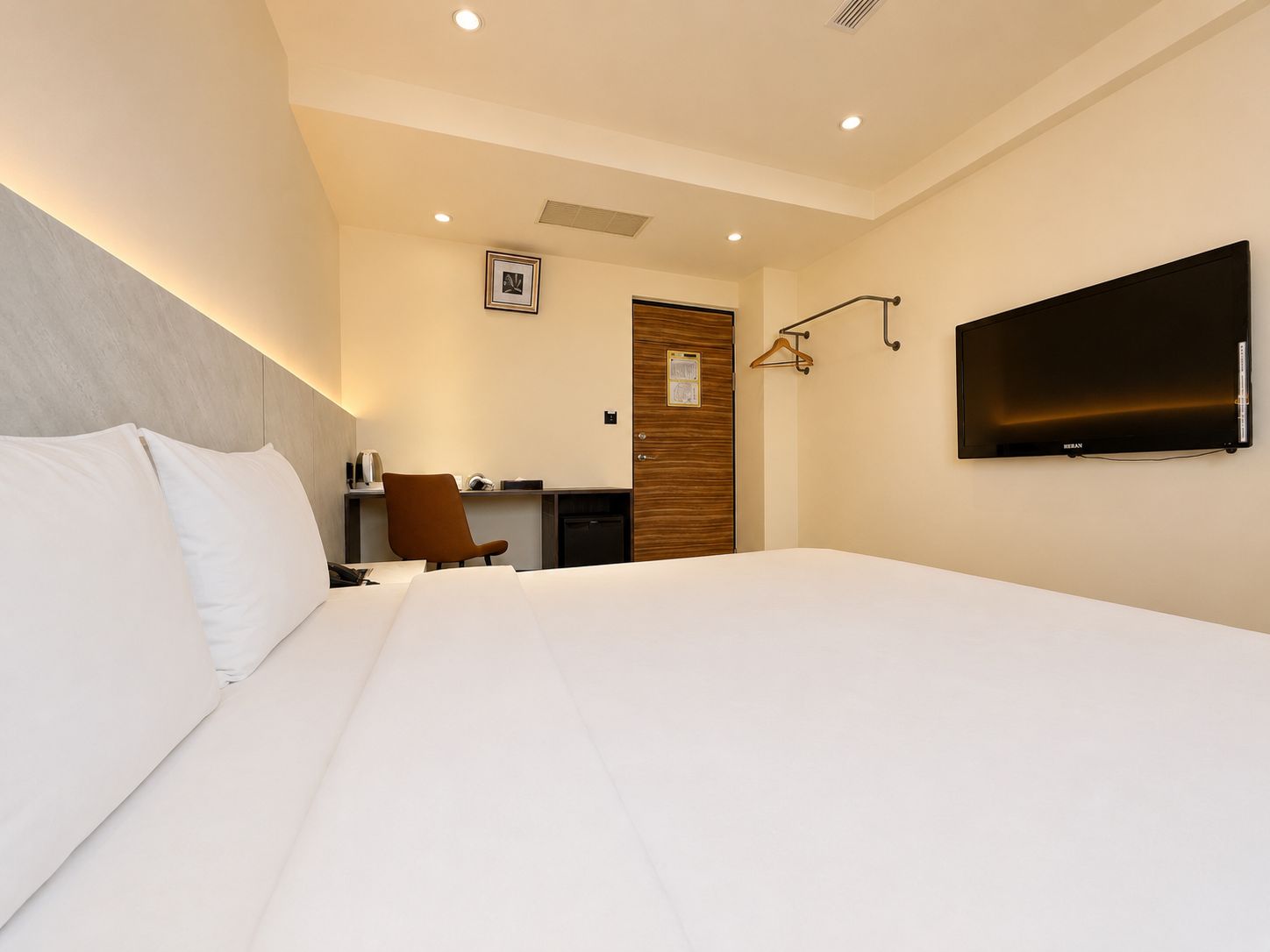 Quarto de hotel com uma cama grande, uma escrivaninha com cadeira, televisão e uma porta com porta-casacos.