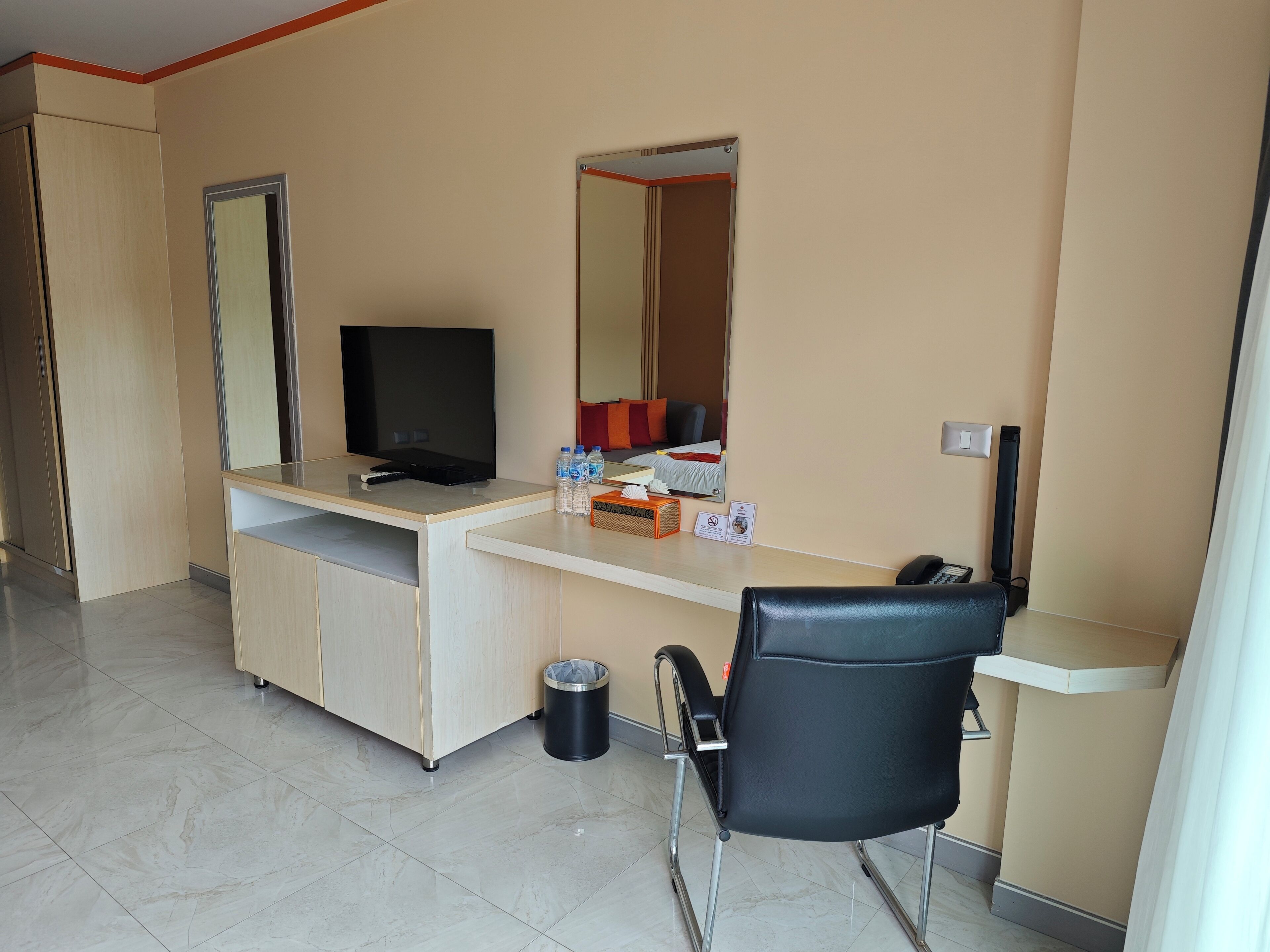 Quarto de hotel com uma escrivaninha de madeira, televisão, espelho, cadeira preta, lixeira e uma pequena mesa de cabeceira com abajur.