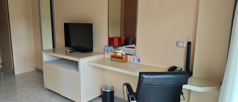 Quarto de hotel com uma escrivaninha de madeira, televisão, espelho, cadeira preta, lixeira e uma pequena mesa de cabeceira com abajur.