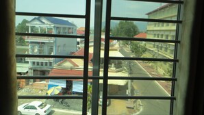 Visão pela janela com grades de metal, mostrando uma rua com edifícios, um carro estacionado e uma motocicleta.
