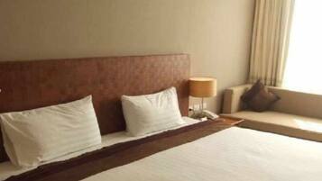 Quarto de hotel com uma cama grande, cabeceira de madeira, mesa de cabeceira com abajur, sofá e janela com cortinas.