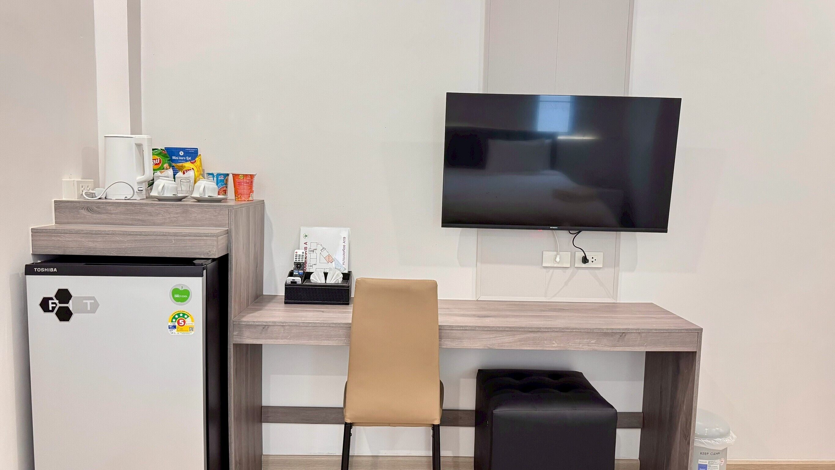 Uma cozinha compacta com geladeira, micro-ondas e cafeteira. Uma TV de tela plana está fixada na parede acima de uma escrivaninha de madeira com uma cadeira.