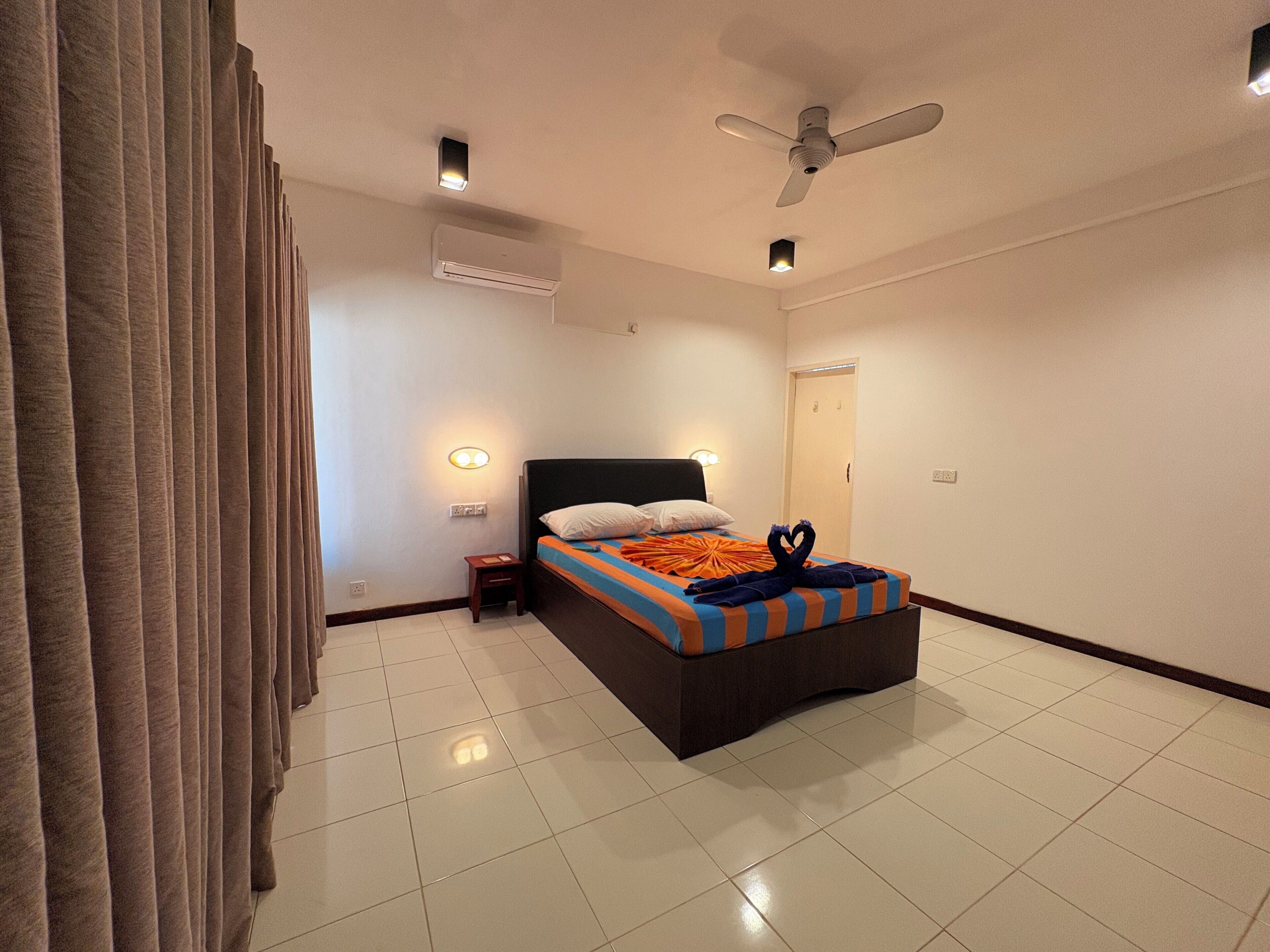 Quarto com cama, ventilador de teto, ar-condicionado e mesa de cabeceira com abajur.