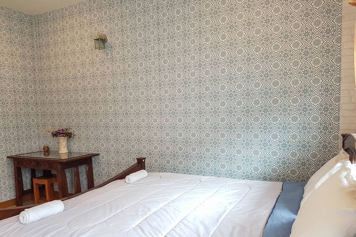 Cama bem arrumada com edredom branco, mesa de cabeceira de madeira com um vaso de flores e parede com padrão.