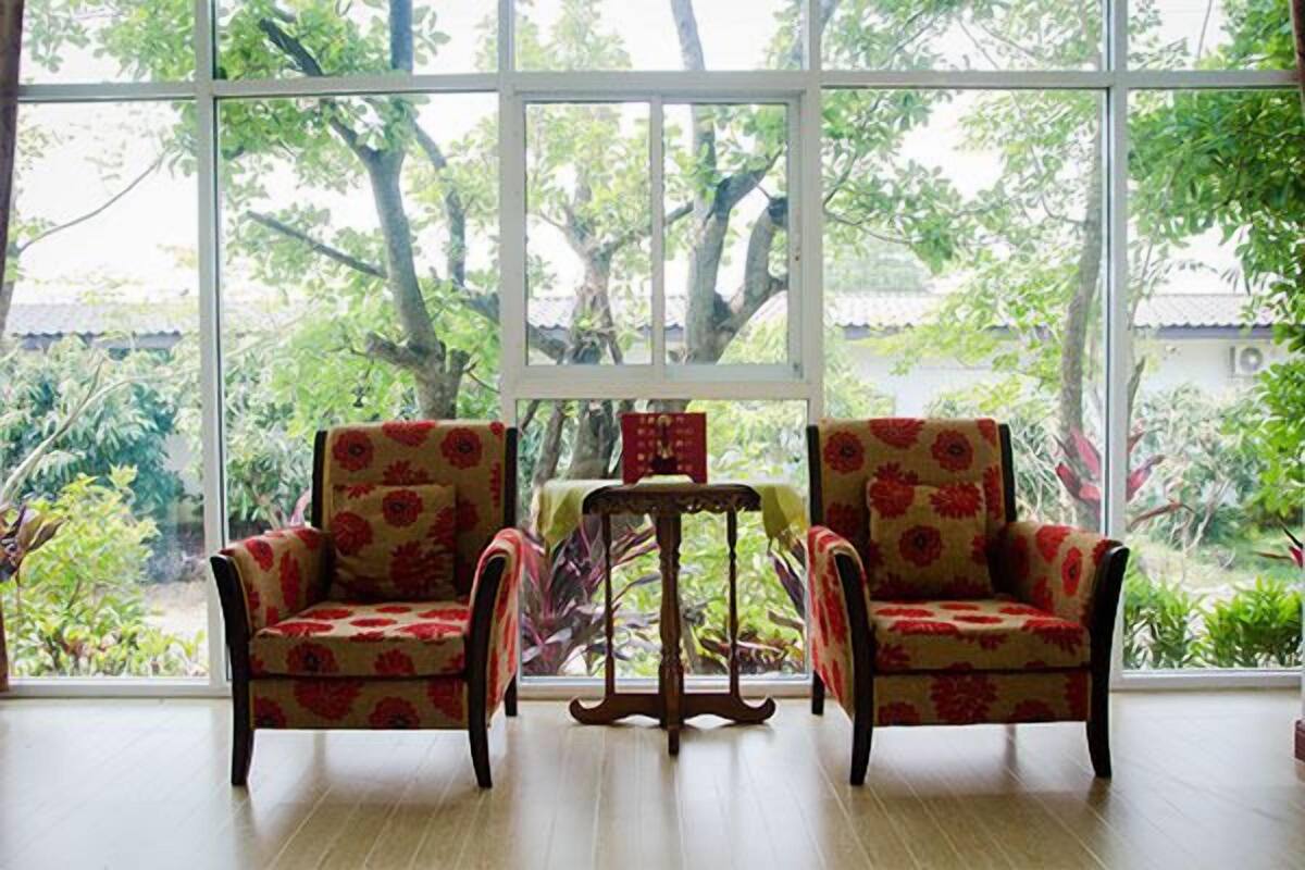 Quarto com duas cadeiras, uma pequena mesa, janelas amplas e vista para árvores e um edifício lá fora.