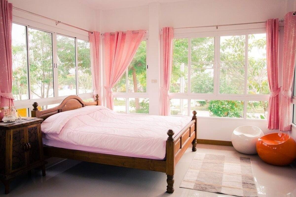 Quarto com cama de madeira, mesa de cabeceira, janela com cortinas rosa e vista para o verde lá fora.