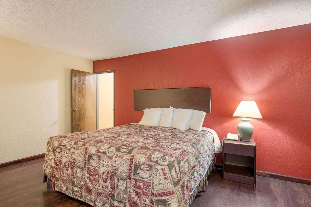 Quarto com uma cama grande, mesa de cabeceira com abajur e porta para outro espaço.