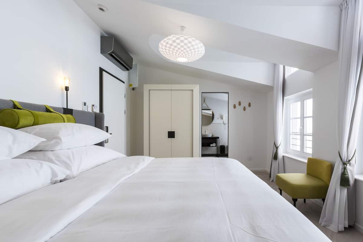 Quarto de hotel moderno com uma cama grande, uma poltrona pequena, um guarda-roupa e uma janela com cortinas.
