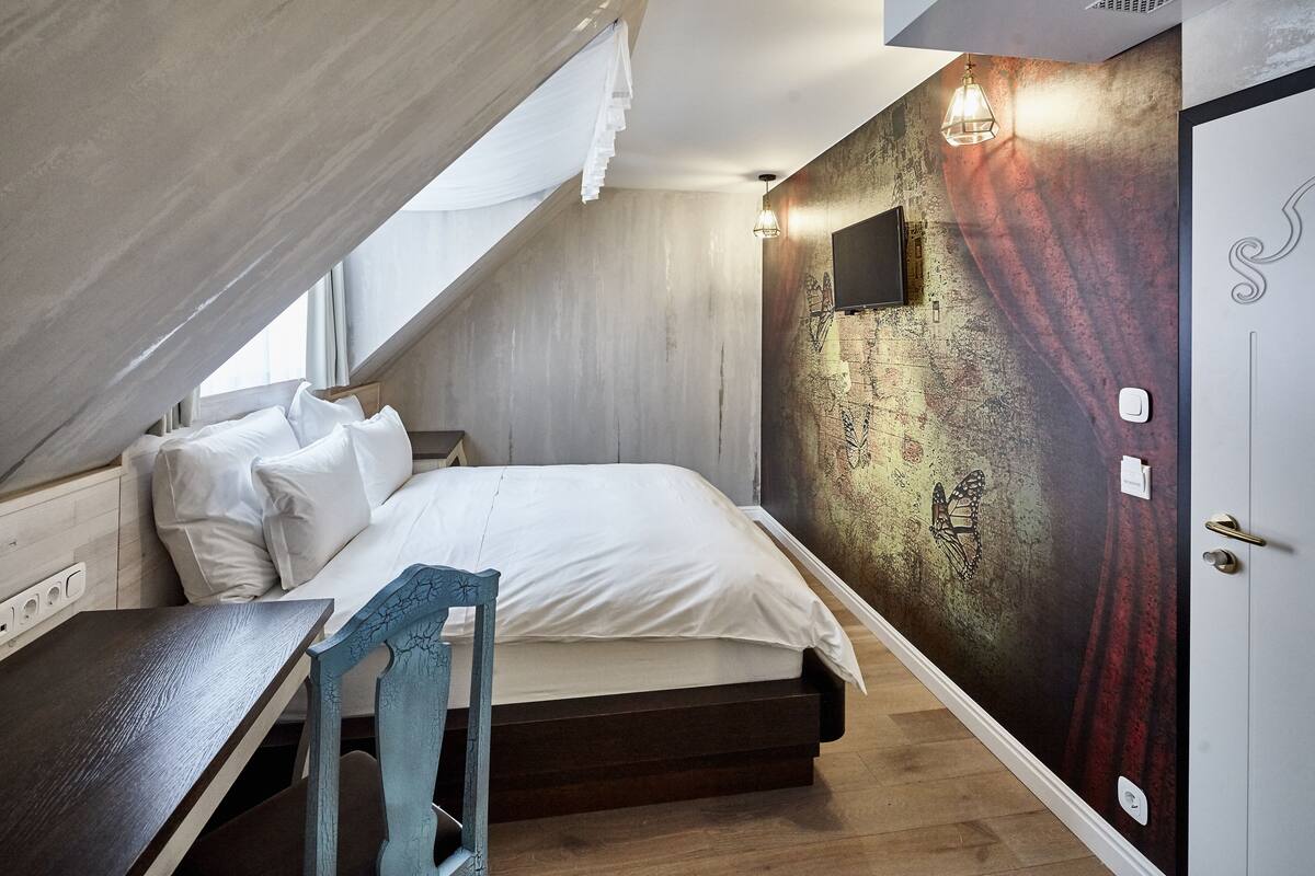 Quarto com cama, escrivaninha e cadeira. Há uma janela com cortinas, uma parede com mural texturizado e uma porta com puxador.