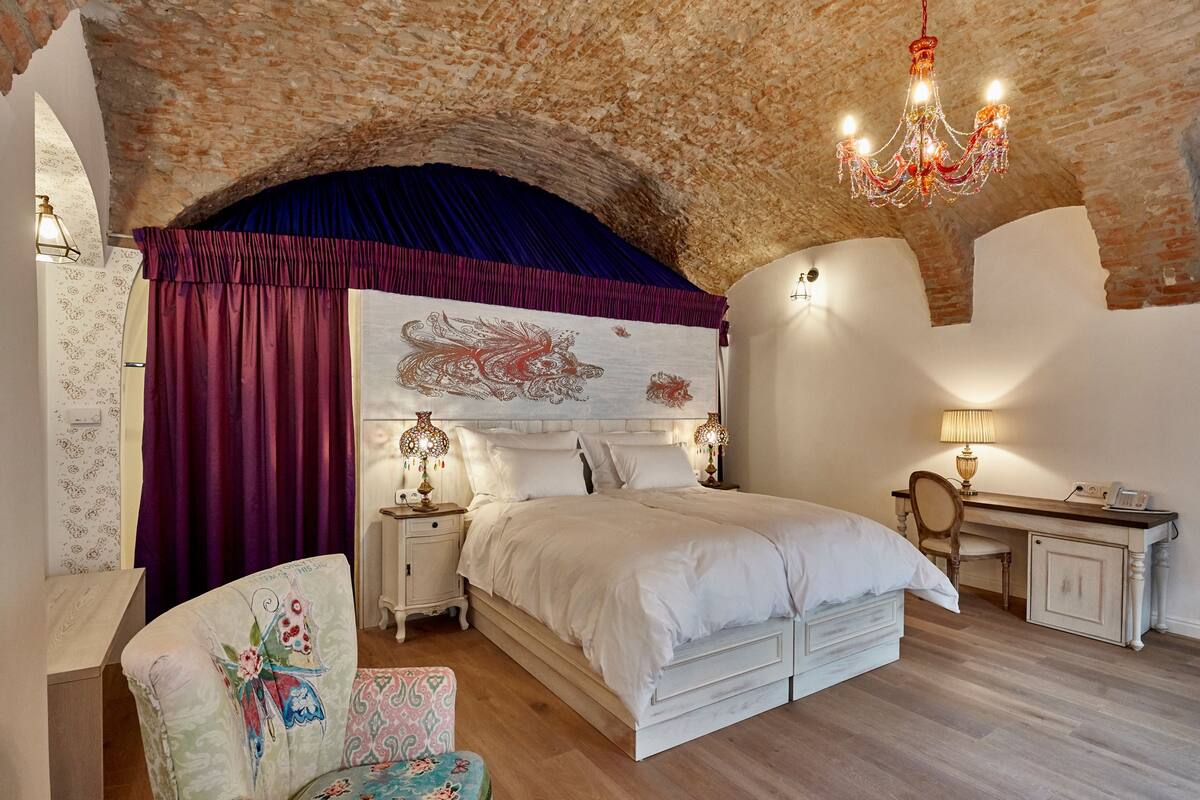 Quarto com uma cama grande, uma escrivaninha e uma cadeira. O quarto possui teto rústico e um lustre.