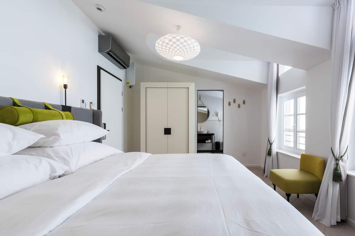 Quarto de hotel moderno com uma cama grande, uma poltrona pequena, um guarda-roupa e uma janela com cortinas.