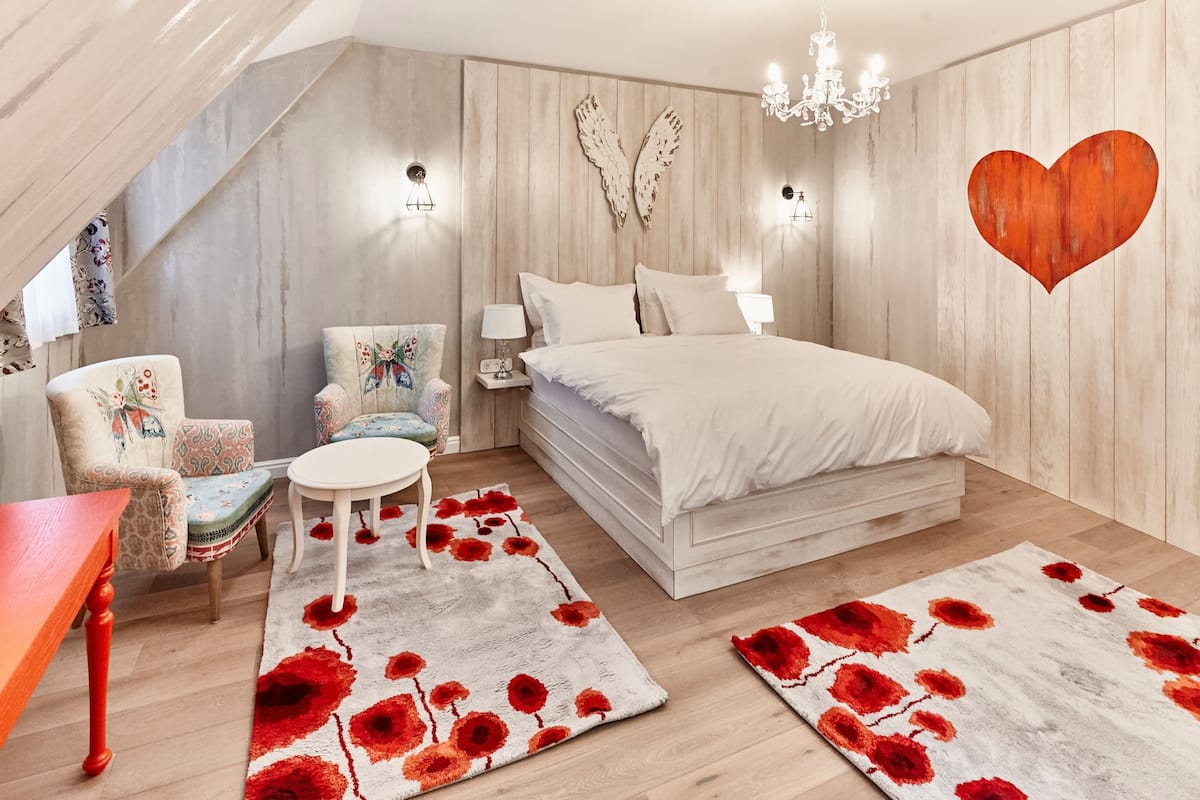 Quarto com uma cama grande, duas mesas de cabeceira, um candelabro, uma decoração em formato de coração e uma poltrona com padrão floral.
