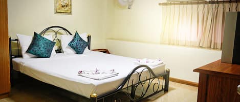 Cama bem arrumada com lençol branco e almofadas estampadas em azul, cabeceira de madeira, mesa de cabeceira pequena, ar condicionado fixado na parede e janela com cortinas bege.